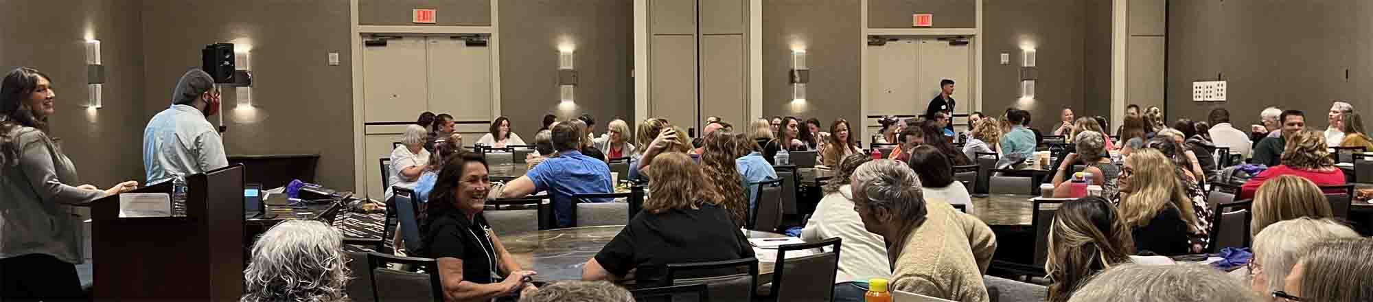picture shows a large room with round tables, several presenters are ...
